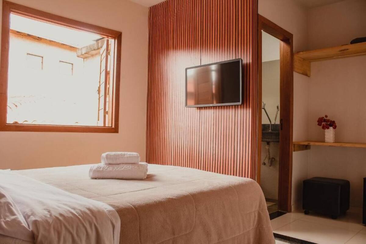 Quarto de hotel com cama, televisão fixada na parede, janela e prateleira de madeira.