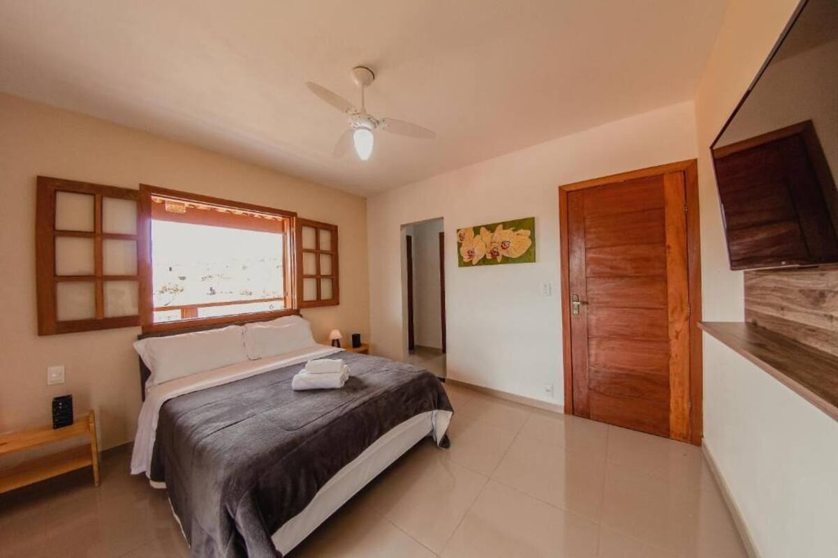 Quarto com cama, ventilador de teto, janela com persianas, porta de madeira e um pequeno banco.