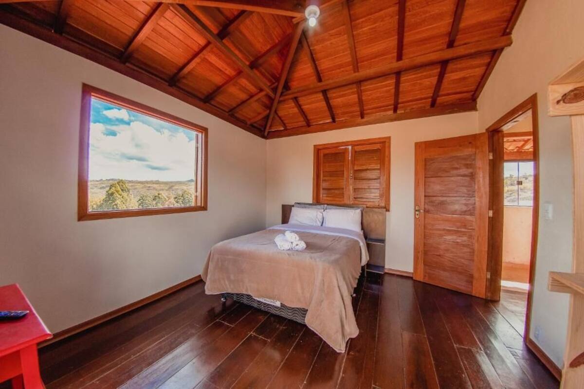 Quarto com teto de madeira, uma cama com cobertura bege, uma janela com vista para a paisagem e uma porta de madeira.