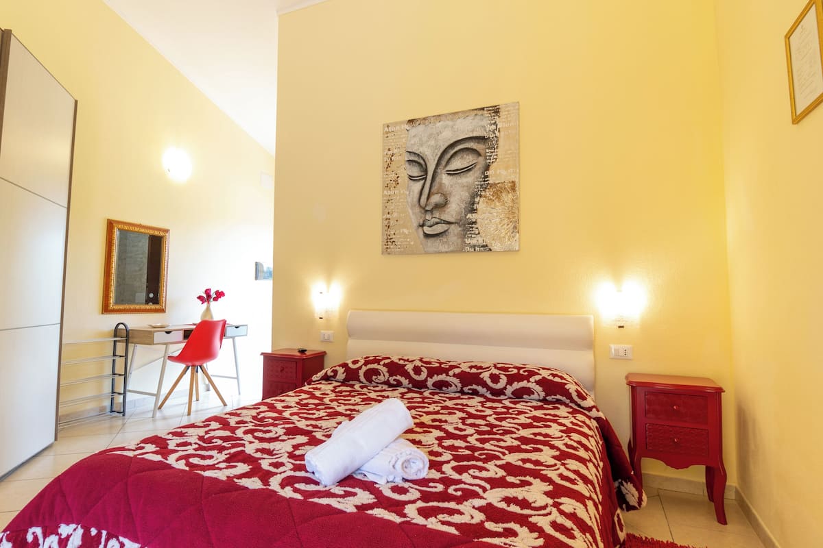 Quarto com cama, escrivaninha, cadeira, espelho e uma obra de arte na parede.