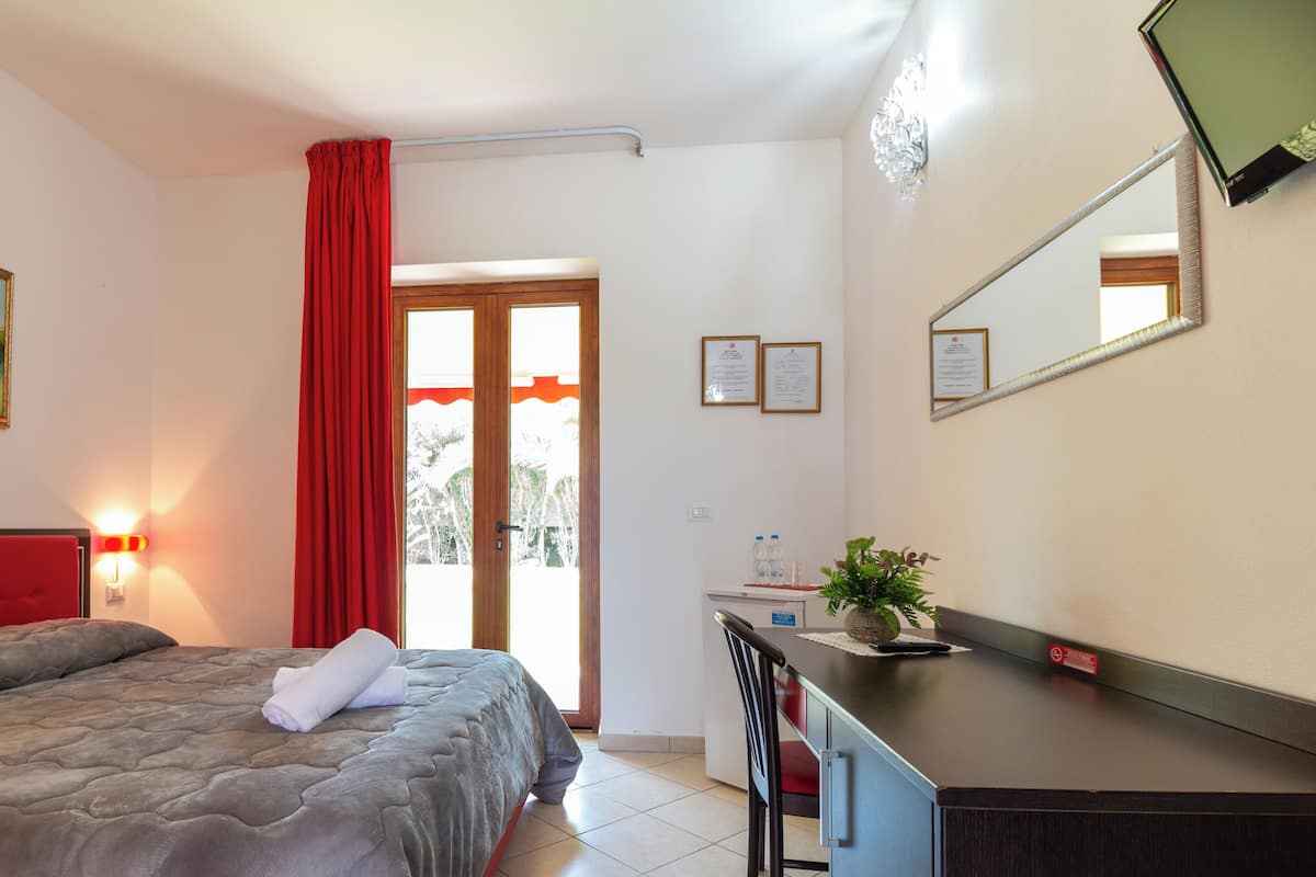 Quarto de hotel com cama, escrivaninha, cadeira, cortina vermelha, quadro e espelho.