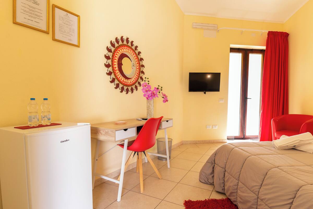 Quarto de hotel com cama, escrivaninha, cadeira, frigorífico, televisão e janela com cortinas.