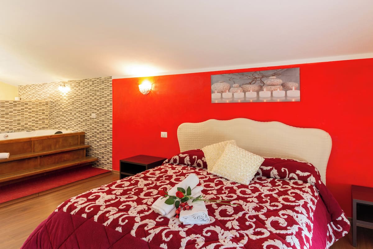 Quarto com parede vermelha, cama grande com cabeceira branca, um banquinho de madeira, colcha com padrão vermelho e branco e um buquê de flores.