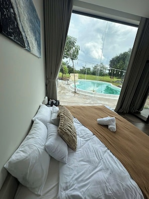 Quarto com cama, travesseiros, toalha e vista para uma piscina.