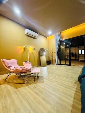 Quarto com uma cadeira rosa, uma pequena mesa, um aparelho de ar condicionado e uma porta de vidro que dá para o exterior.