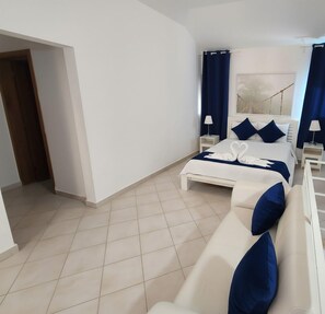 Quarto de hotel com cama, duas mesas de cabeceira, um criado-mudo, um sofá e uma porta que dá para outro cômodo.