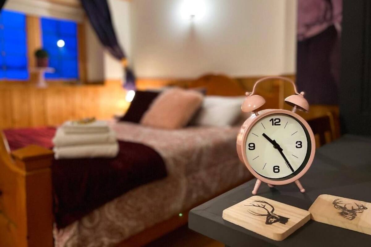 Uma cama com cabeceira, uma mesa de cabeceira com um relógio e uma pequena mesa com duas placas de madeira.