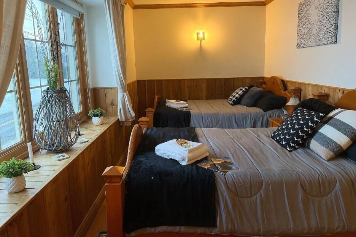Quarto de hotel com duas camas, uma mesa de cabeceira, abajur, luz embutida na parede e janela com cortinas.