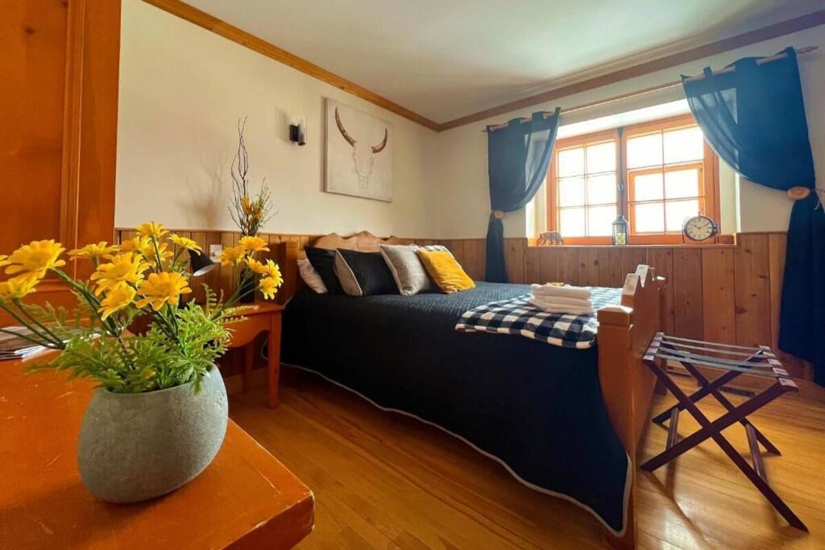 Um quarto com uma cama, uma cadeira de madeira, uma mesa com um vaso de flores, uma janela com cortinas e um relógio de parede.