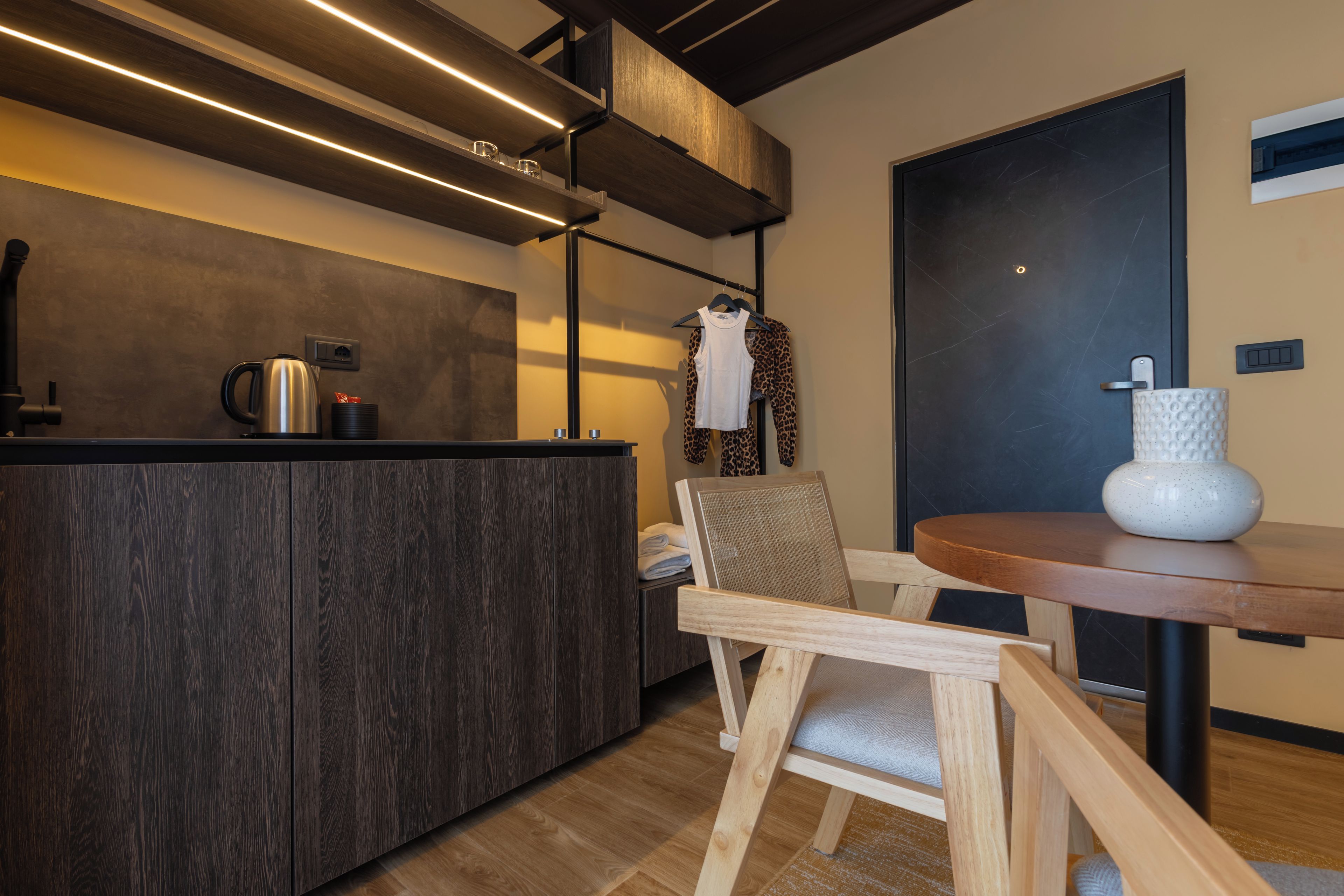 Cozinha moderna com bancada de madeira escura, chaleira e máquina de café. Há uma cadeira com assento trançado e uma pequena mesa com um vaso branco.