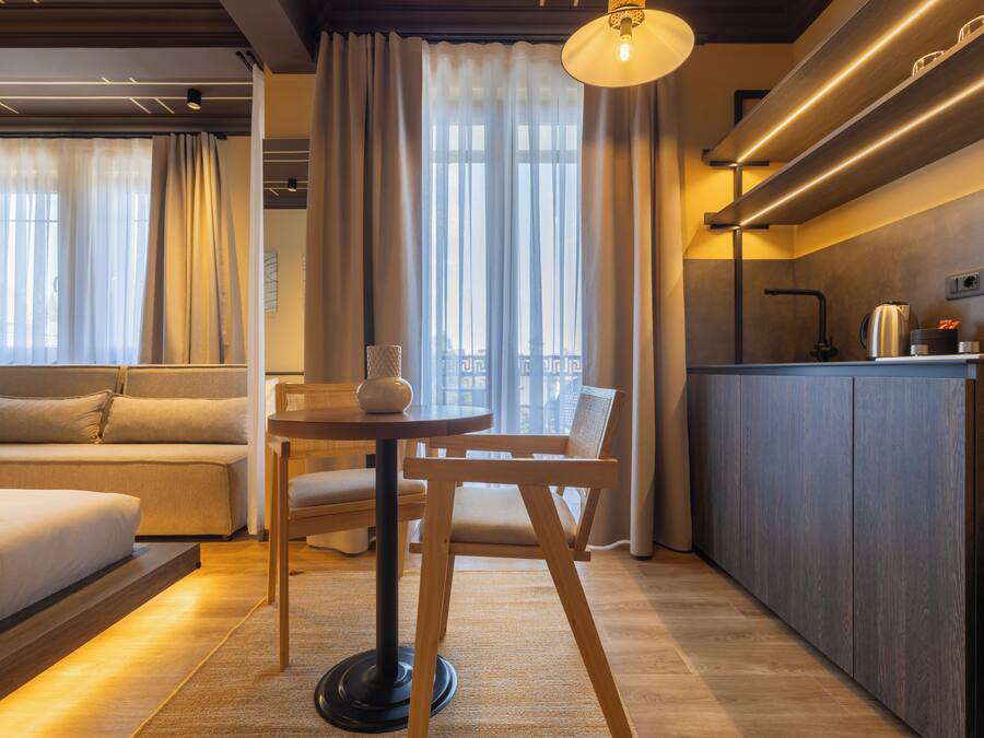 Quarto de hotel moderno com sofá, mesa redonda pequena, uma cadeira, área de cozinha com bancada e armários, e janelas amplas com cortinas.