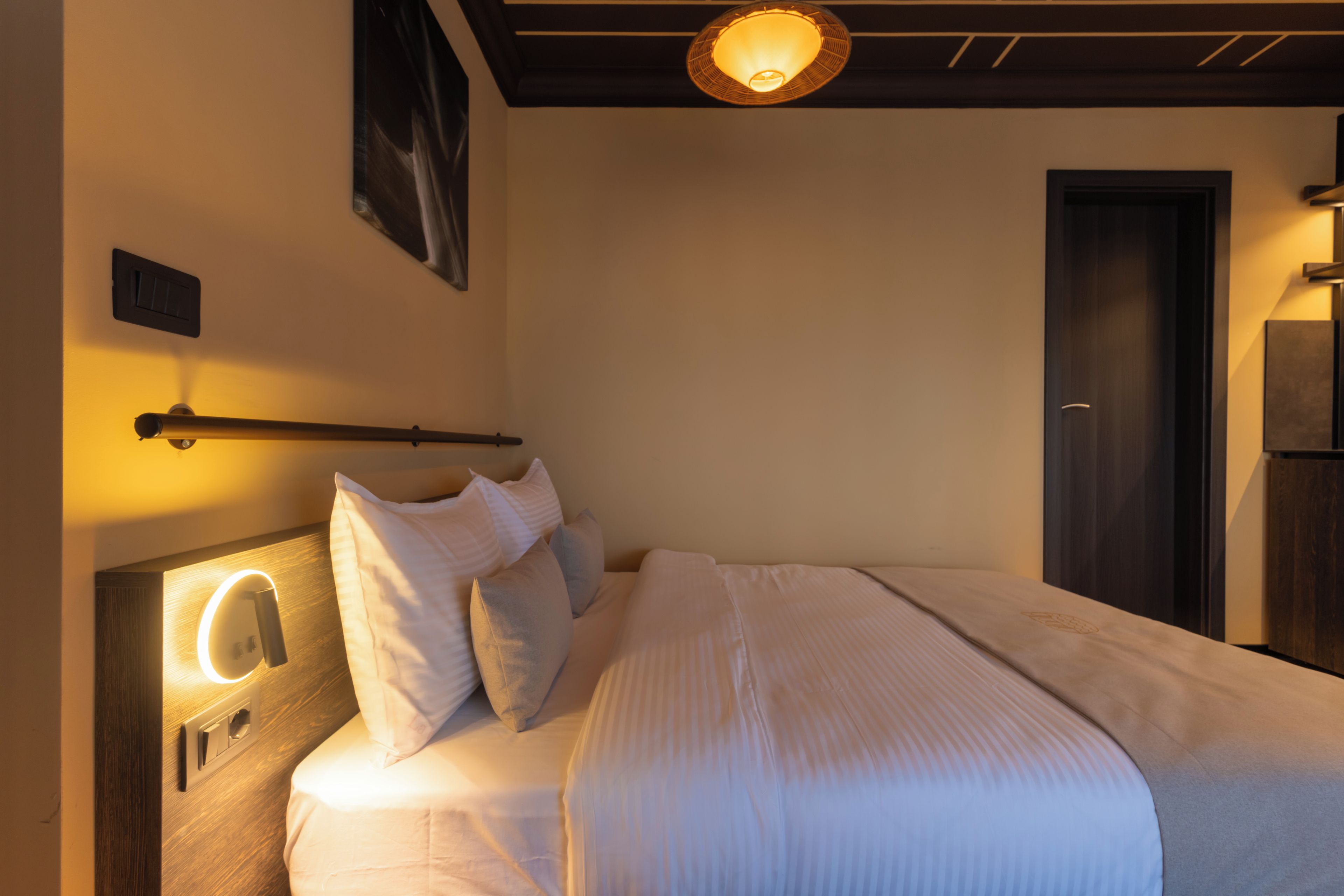 Quarto de hotel com cama, controles laterais, luminária na parede e uma porta.