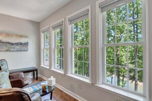 Quarto com grandes janelas, um banco, uma mesa com um livro e uma xícara, e um quadro na parede.