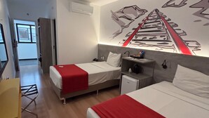 Quarto de hotel com duas camas, uma escrivaninha, uma cadeira, um pequeno frigorífico e um mural na parede.