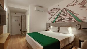 Quarto de hotel com cama, escrivaninha, cadeira, luminária na parede e porta para outro cômodo.