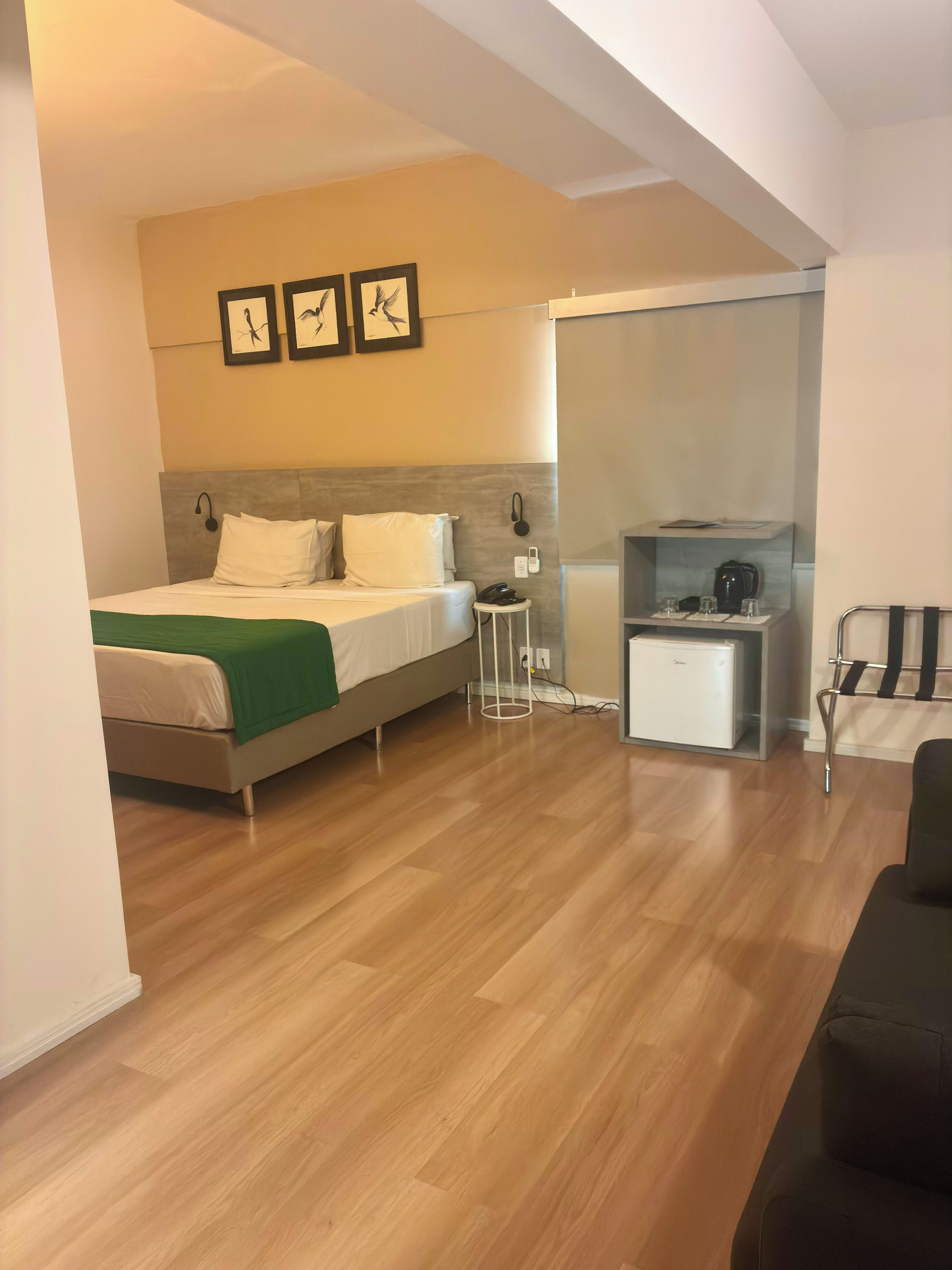 Quarto de hotel com cama, mesa de cabeceira, pequena escrivaninha com cadeira, máquina de café e porta-roupas.