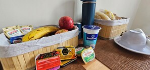 Mesa de café da manhã com cesta contendo uma banana, uma maçã e pão, uma garrafa térmica, um copo de iogurte e um prato com uma chaleira.