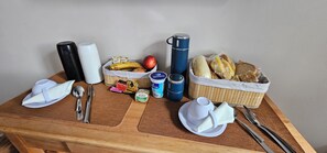 Disposição para o café da manhã com pão, frutas, acompanhamentos e uma garrafa térmica em uma mesa de madeira.