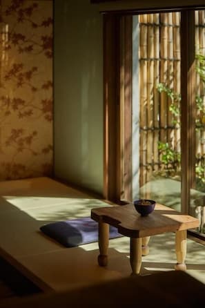 Um quarto com área de estar com esteira de tatame, uma pequena mesa de madeira com uma tigela e um biombô de bambu.