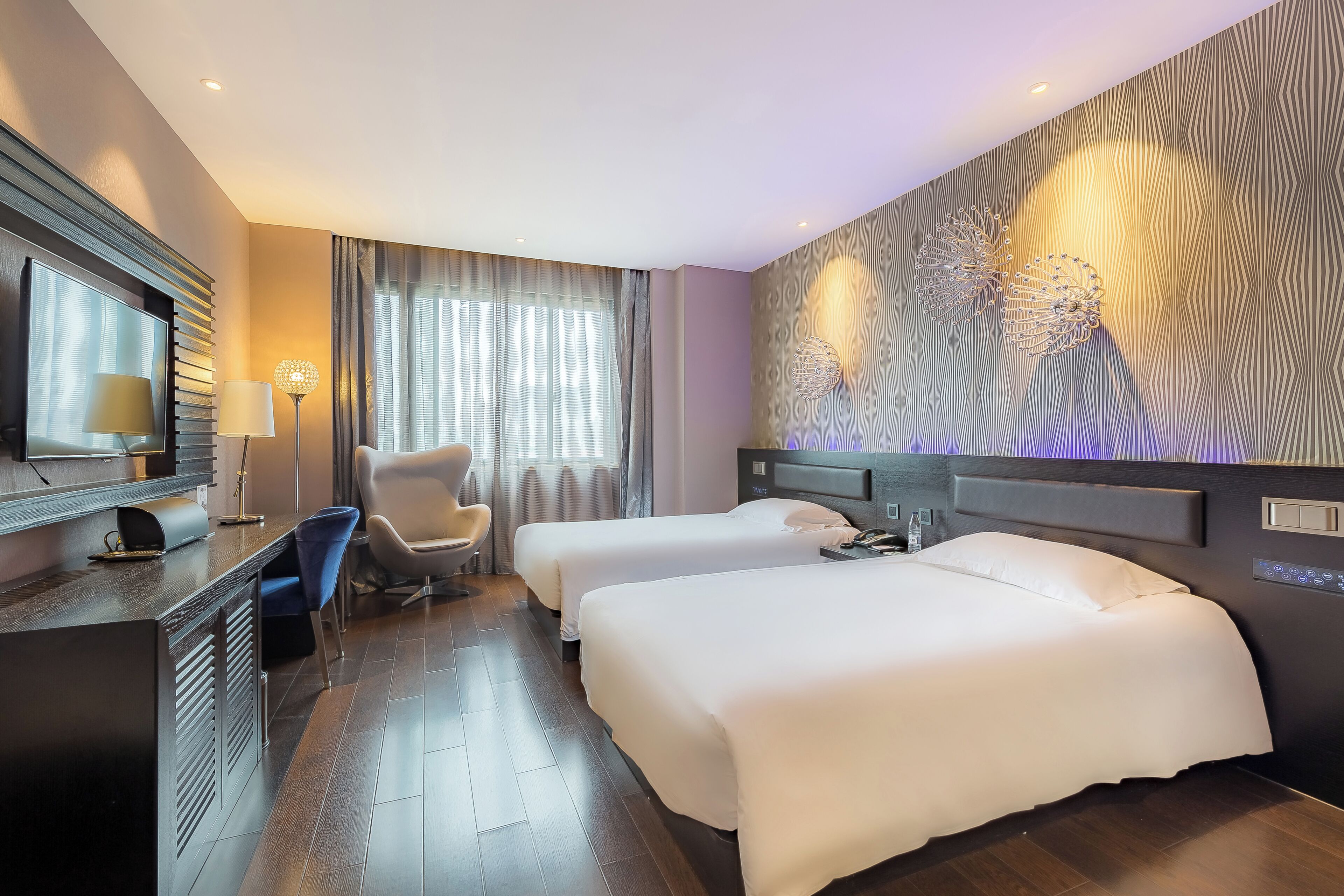 Quarto de hotel com duas camas, uma escrivaninha com televisão, uma cadeira e um painel decorativo.