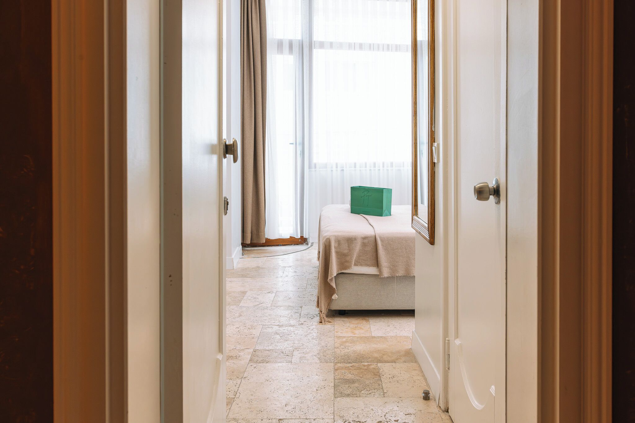 Quarto de hotel com uma cama, uma caixa verde e uma janela com cortinas leves.
