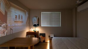Quarto de hotel com cama, escrivaninha, cadeira, luminária, janela com persianas e tela de projeção acoplada à parede exibindo uma cena de filme.