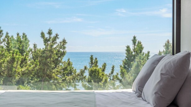 Uma cama com roupa de cama e almofadas brancas, uma janela vista para o mar e árvores.
