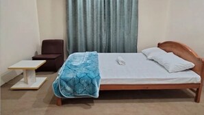 Uma cama de solteiro com cabeceira de madeira, uma mesinha de cabeceira, uma cadeira e uma janela com cortinas.
