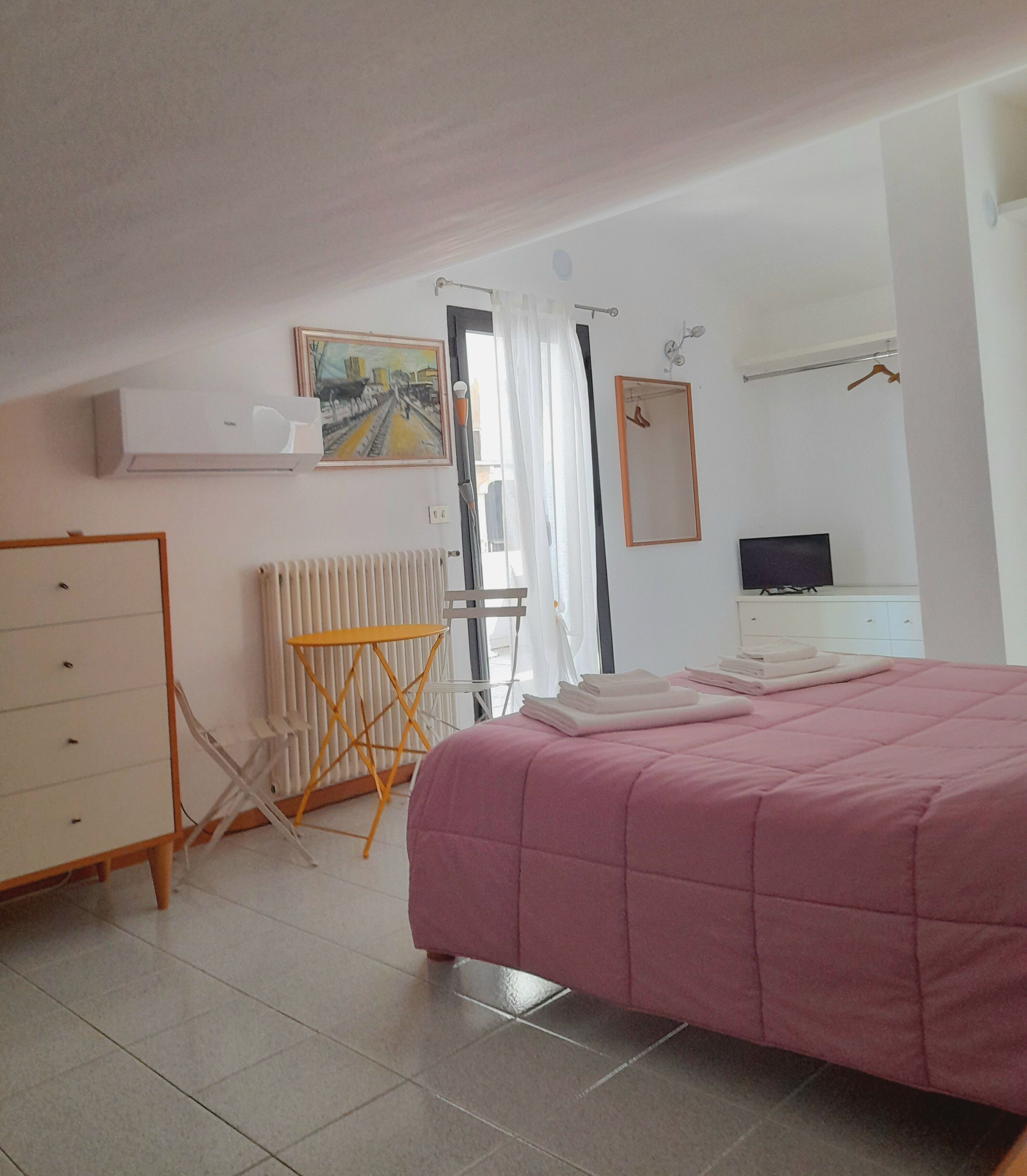 Um quarto com uma cama, um radiador, um varal, uma cômoda, uma televisão e um quadro na parede.