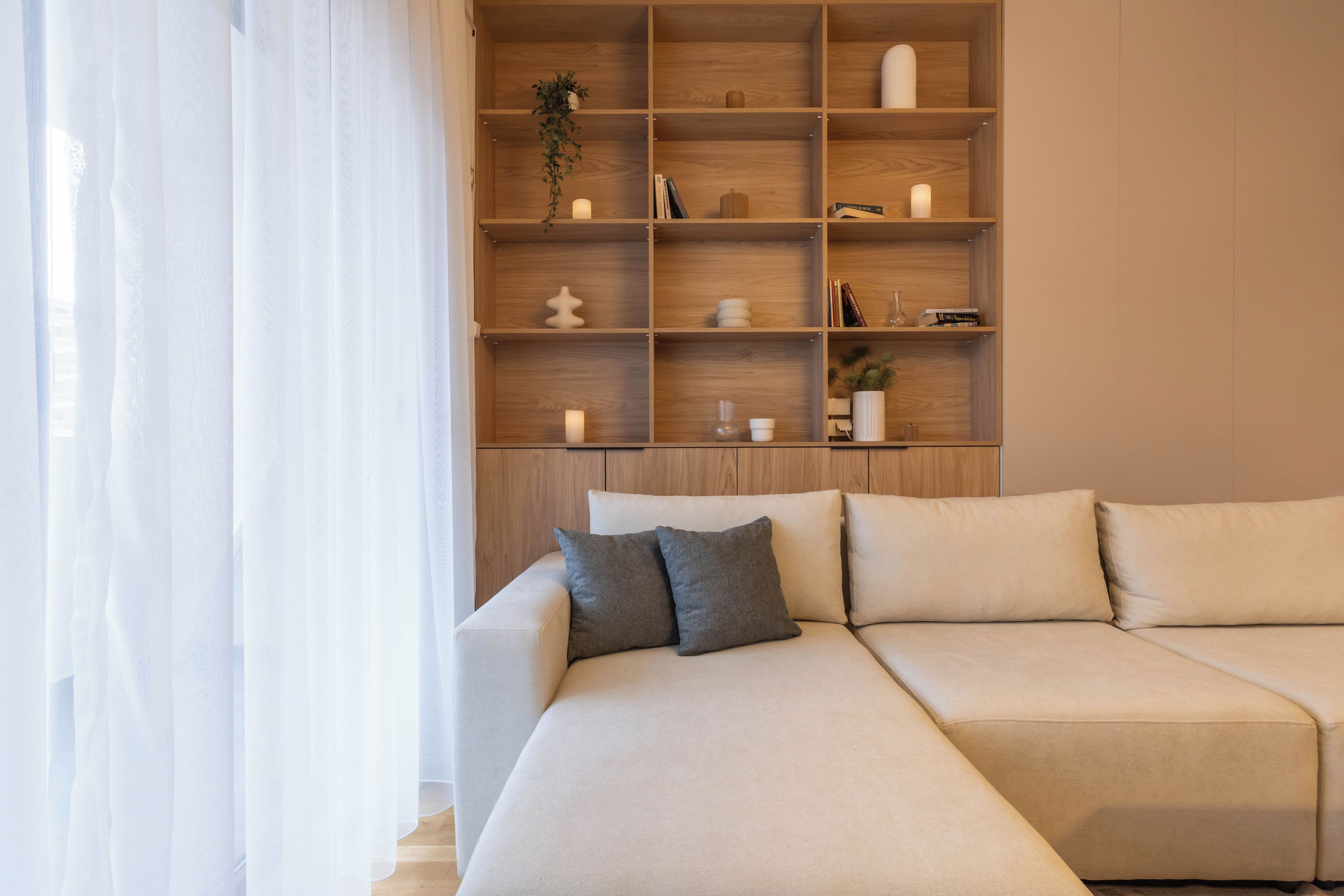 Uma sala de estar moderna com um sofá bege, duas almofadas cinzentas e uma estante de madeira com diversos objetos decorativos.