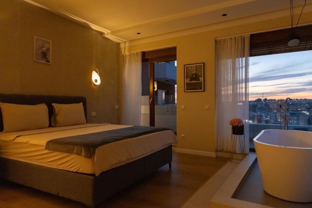 Quarto com cama, banheira, vista para a cidade e porta que dá para uma varanda.