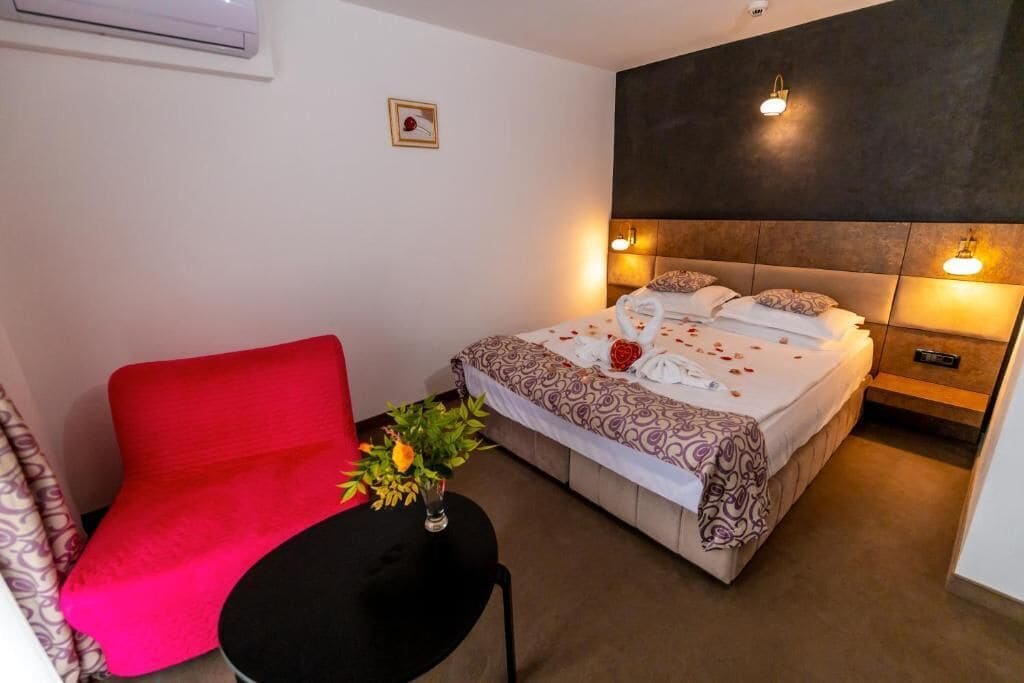 Quarto de hotel com uma cama, uma cadeira vermelha, uma mesinha com um vaso e uma luminária na parede.
