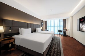 Quarto de hotel com duas camas, uma escrivaninha, uma cadeira, uma TV e uma janela grande.