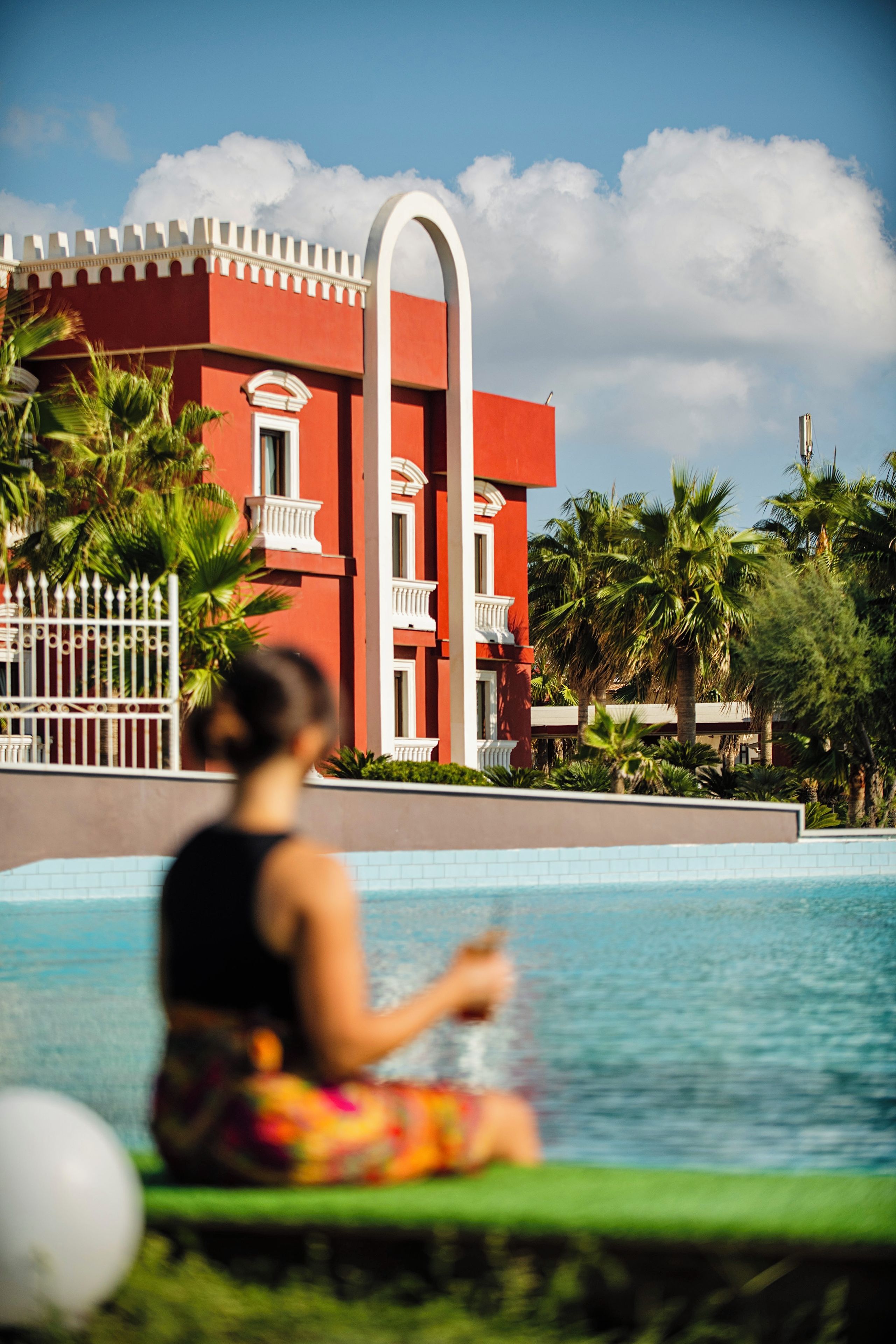 Uma pessoa sentada à beira da piscina, com um edifício vermelho ao fundo.