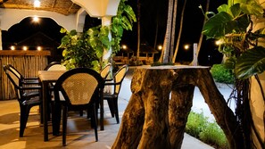 Uma área de refeições ao ar livre com mesa e cadeiras, cercada por plantas e um toco de árvore.