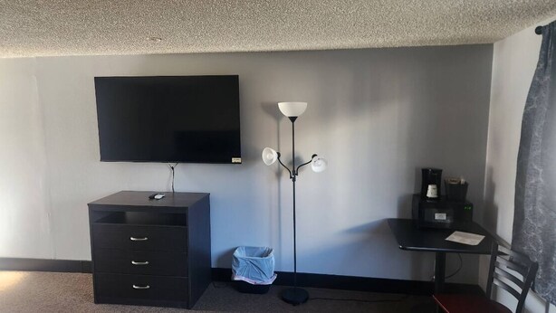 Quarto com televisão de tela plana na parede, escrivaninha preta com cafeteira, luminária de chão e armário de arquivos preto.