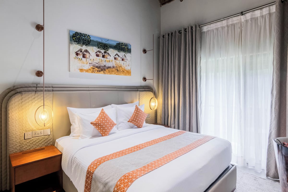 Cama bem arrumada com colcha estampada, mesa de cabeceira com abajur, pintura na parede e janela com cortinas.