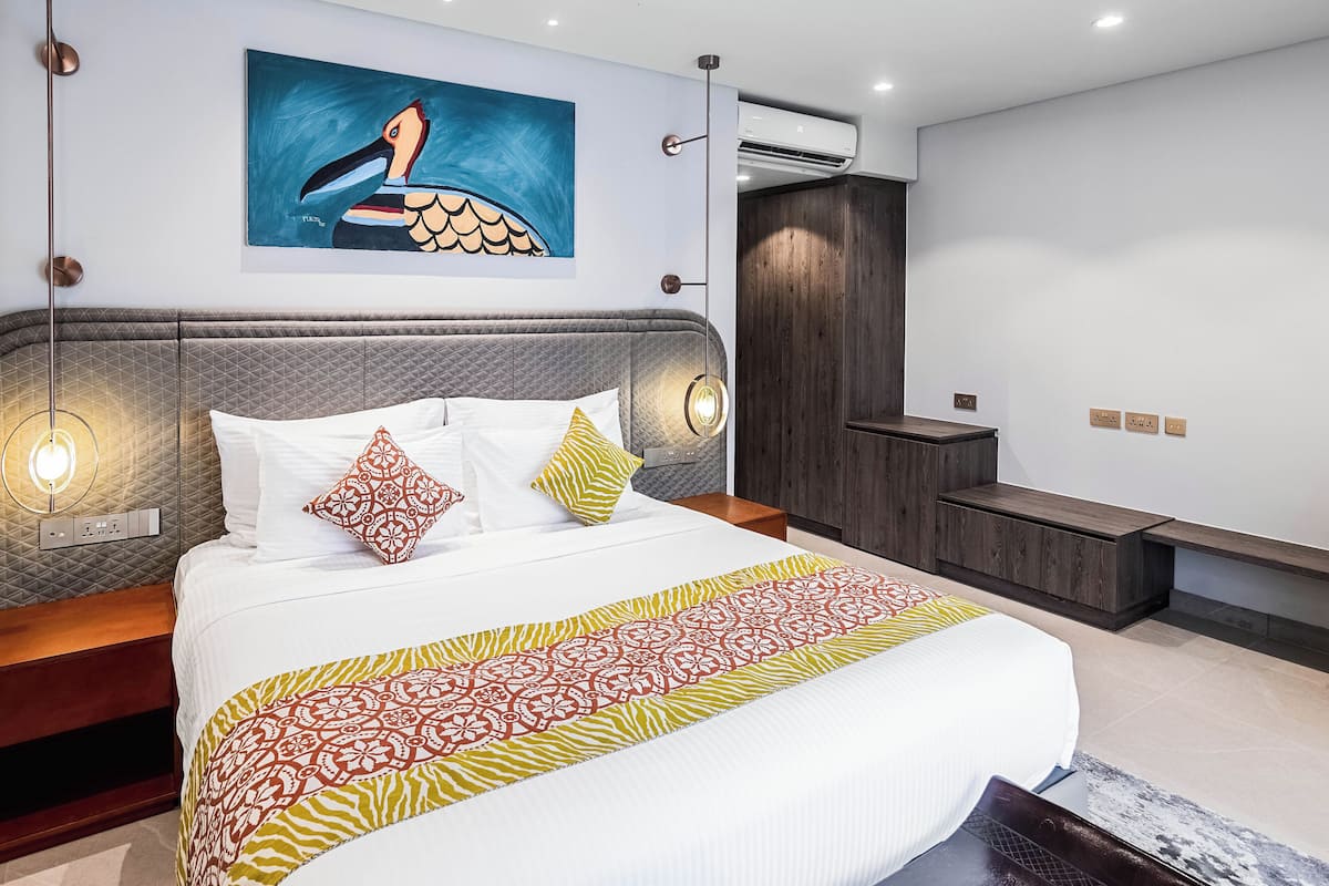 Quarto de hotel moderno com uma cama grande, cabeceira com iluminação embutida, uma obra de arte pendurada na parede e uma mesa de cabeceira de madeira.