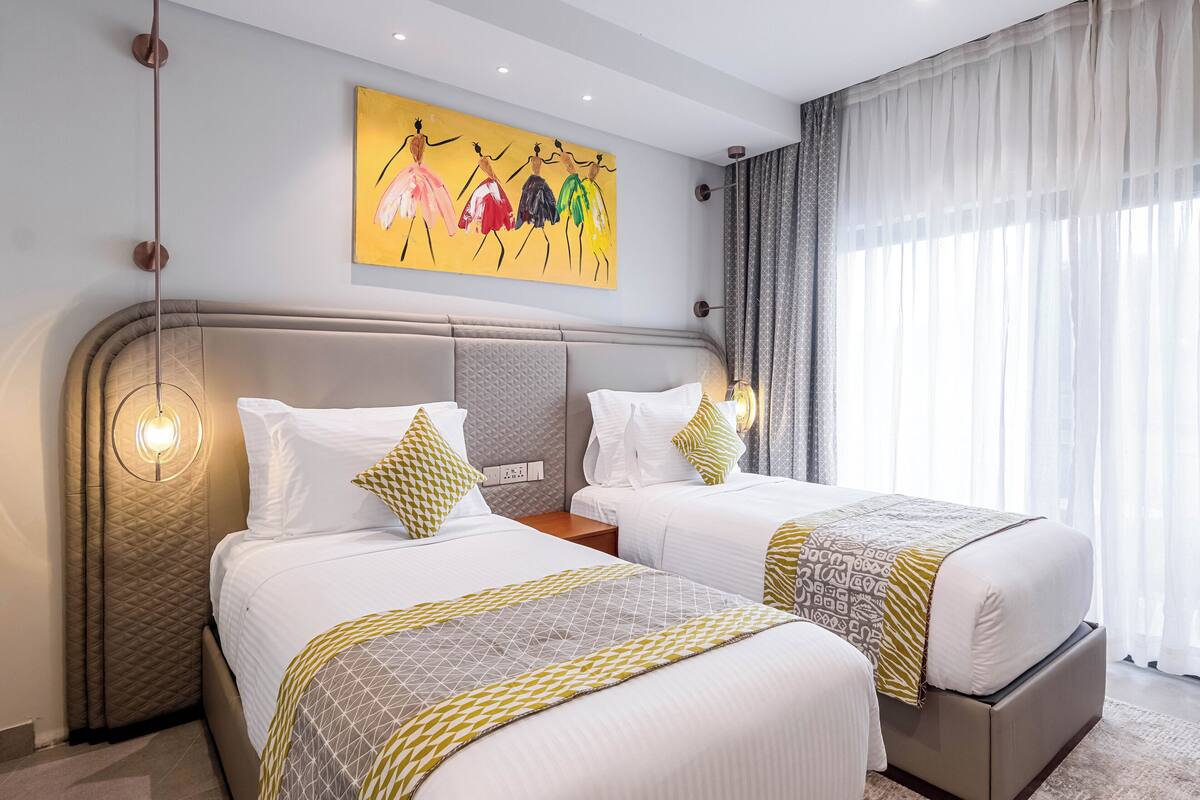 Quarto de hotel com duas camas, uma janela grande com cortinas translúcidas, uma obra de arte na parede e uma luminária moderna.