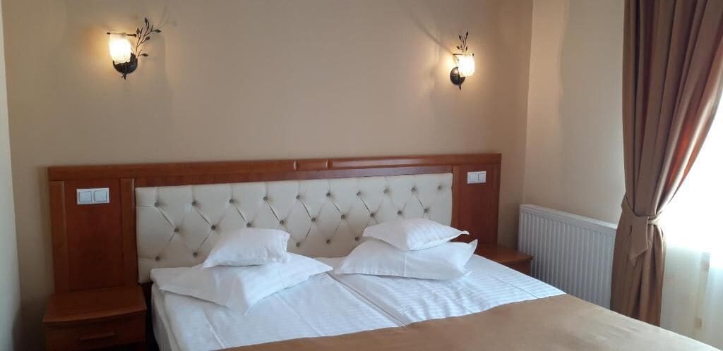 Cama bem arrumada com cabeceira de madeira, duas luminárias fixadas na parede e uma janela com cortinas.