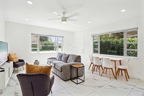 Uma sala de estar moderna com um sofá cinzento, uma pequena mesa de apoio de madeira e uma área de jantar com cadeiras brancas e uma mesa.