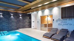 Uma área interna de piscina moderna, com piscina translúcida, vigas de madeira no teto e espreguiçadeiras.