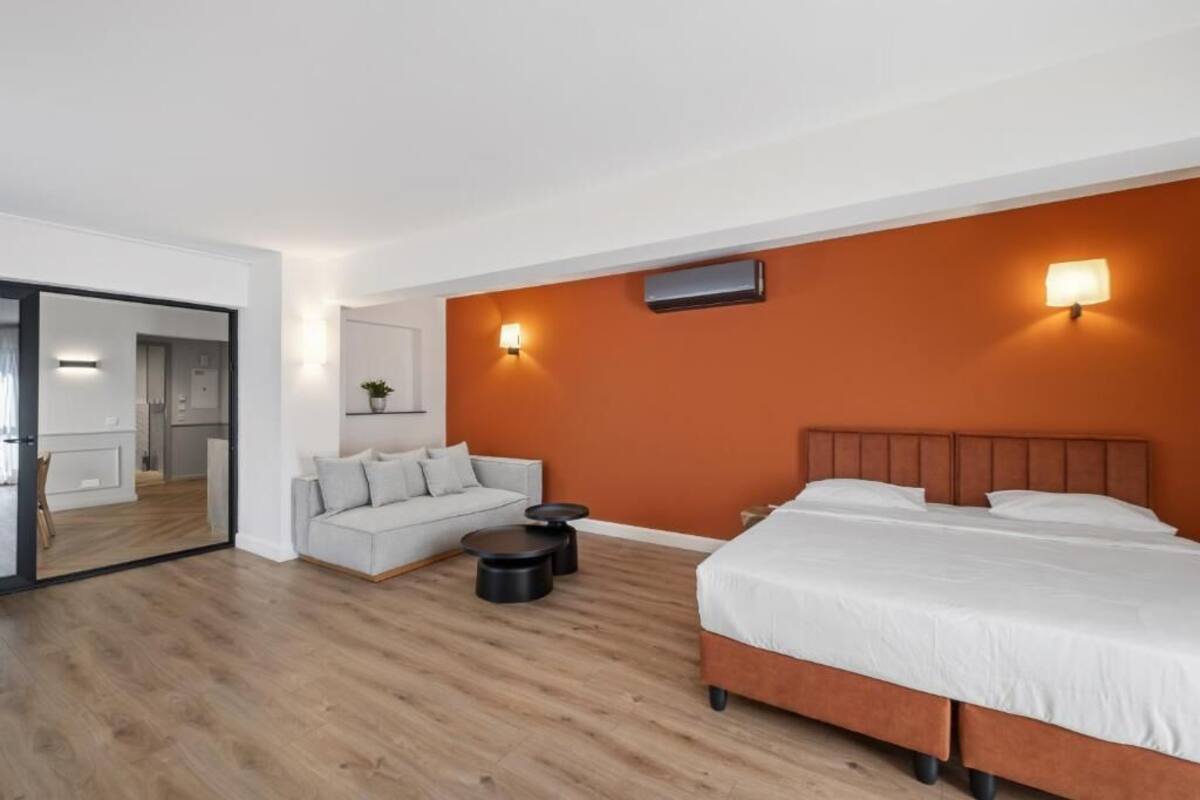 Quarto de hotel moderno com piso de madeira, sofá branco, cama com cabeceira de madeira, duas luminárias fixadas na parede e uma pequena mesa.