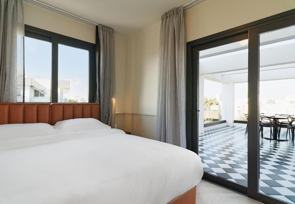 Quarto de hotel com cama, cortinas, vista da cidade e varanda com piso xadrez.