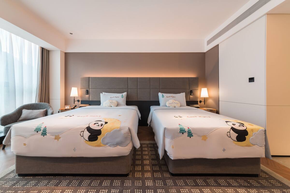 Quarto de hotel com duas camas, um sofá, uma janela grande com cortinas e um tapete estampado.