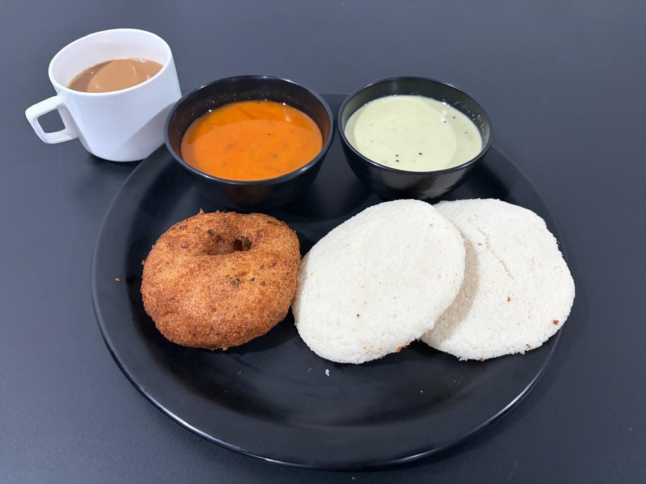 Um prato com dois pães, um alimento frito e duas pequenas tigelas de molho sobre uma superfície escura.