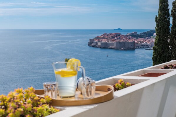 Varanda com uma bandeja contendo um copo de água e um limão, com vista para uma cidade costeira com edifícios históricos e mar azul límpido.