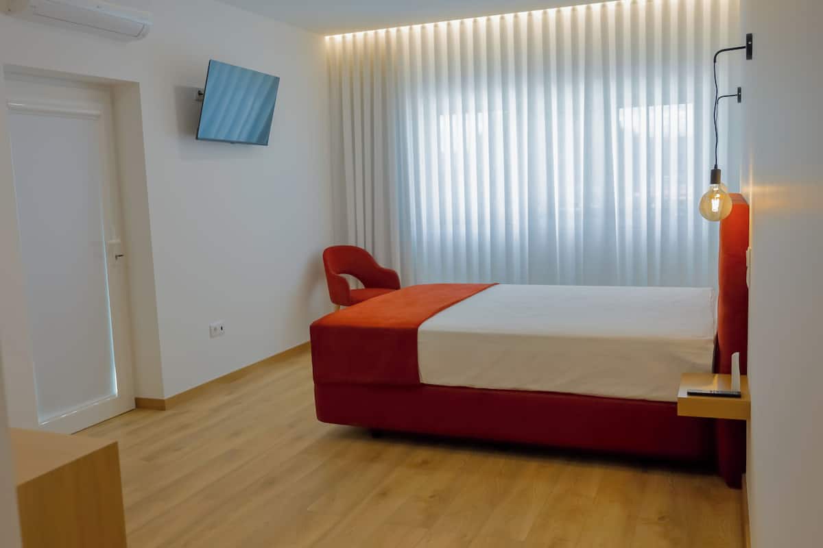 Quarto com cama, cadeira vermelha, mesa de cabeceira, TV na parede e janela com persianas.