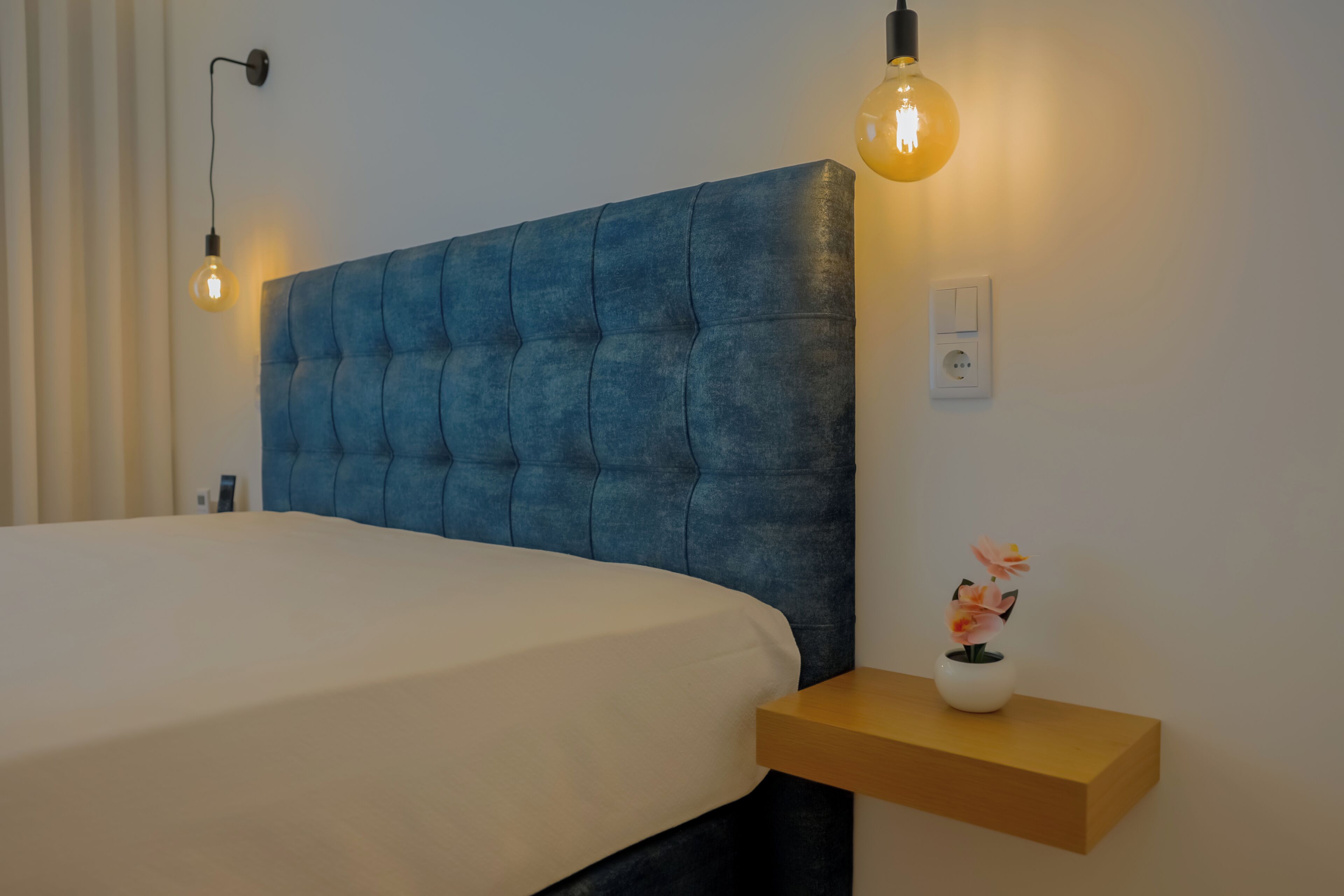 Cama com cabeceira estofada, mesa de cabeceira com um vaso de flores e luminárias fixadas na parede.