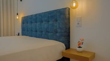 Cama com cabeceira estofada, mesa de cabeceira com um vaso de flores e luminárias fixadas na parede.